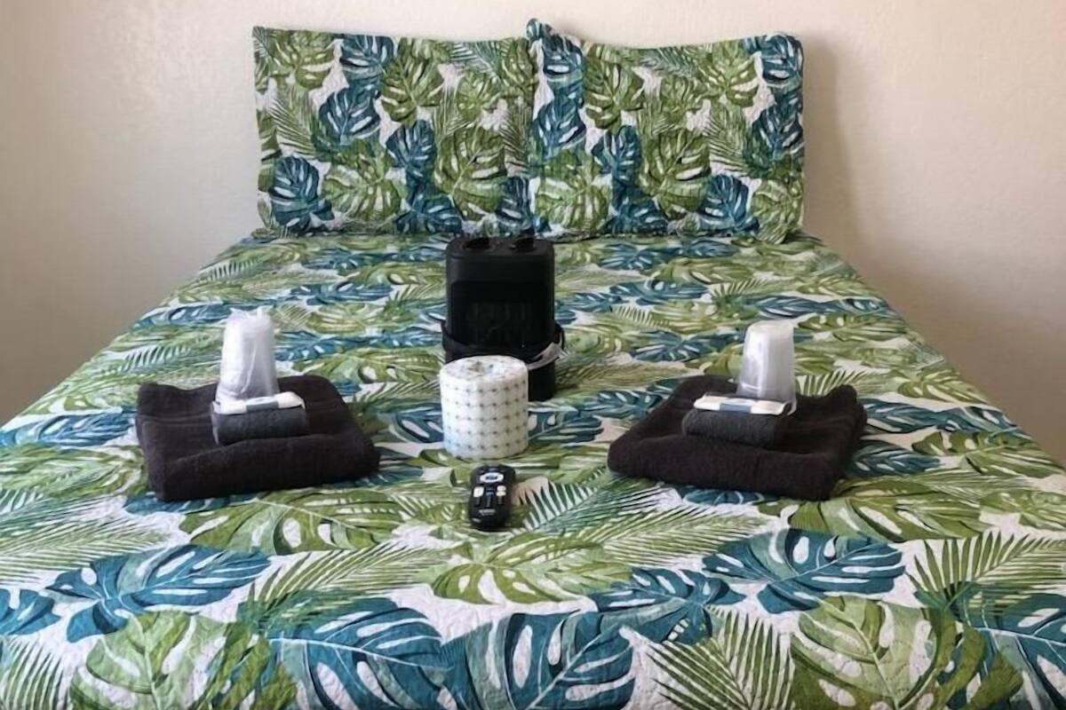 Uma cama com colcha estampada com folhas tropicais, duas toalhas castanho-escuras, um controle remoto preto e um rolo de papel higiênico.