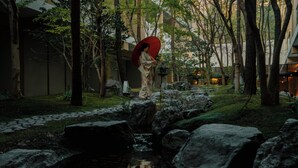 Um jardim japonês tradicional com um caminho de pedra, pedras e uma pessoa segurando um leque vermelho.