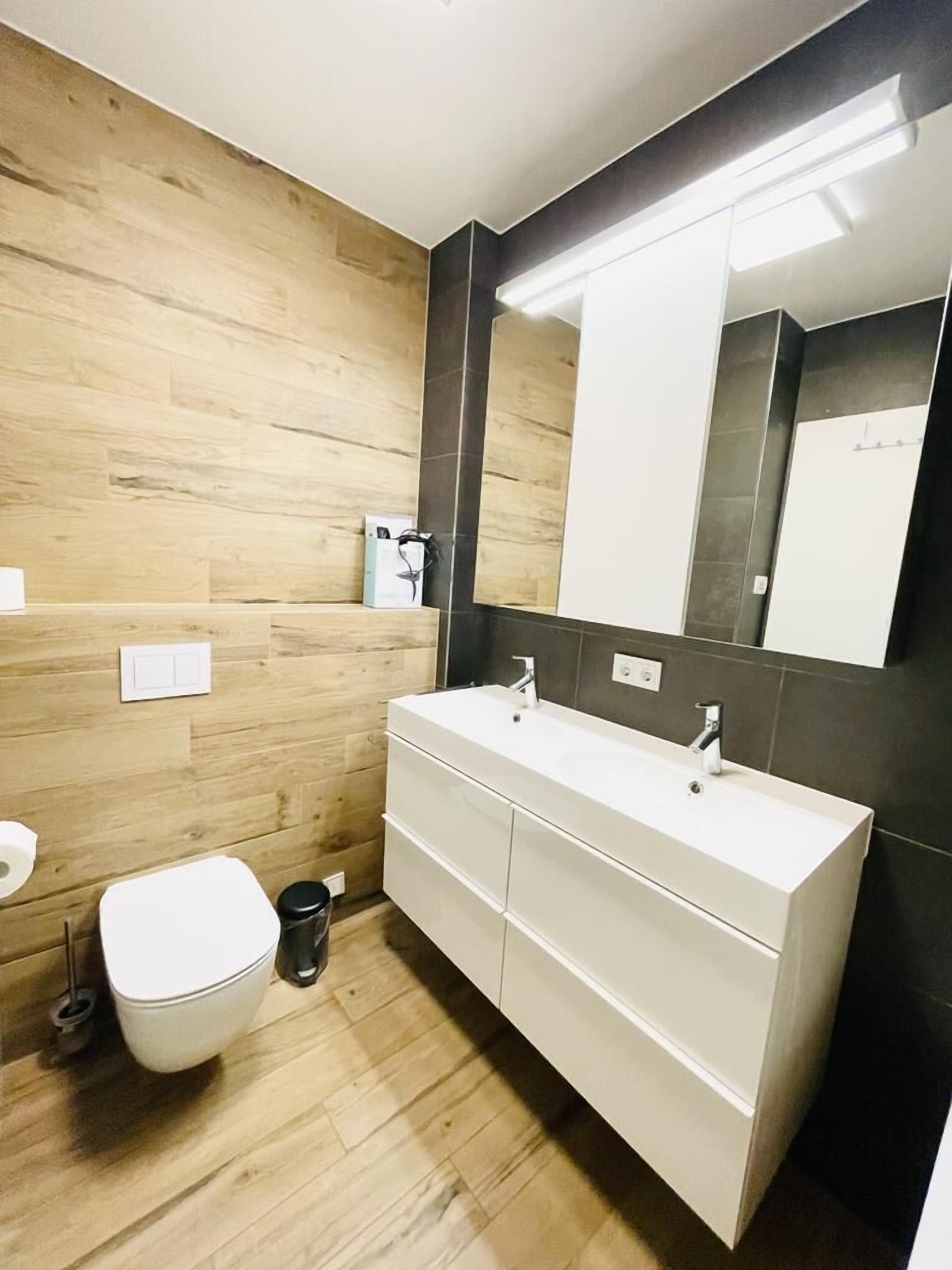 Casa de banho moderna com soalho de madeira, lavatório branco duplo, espelho grande e sanita.