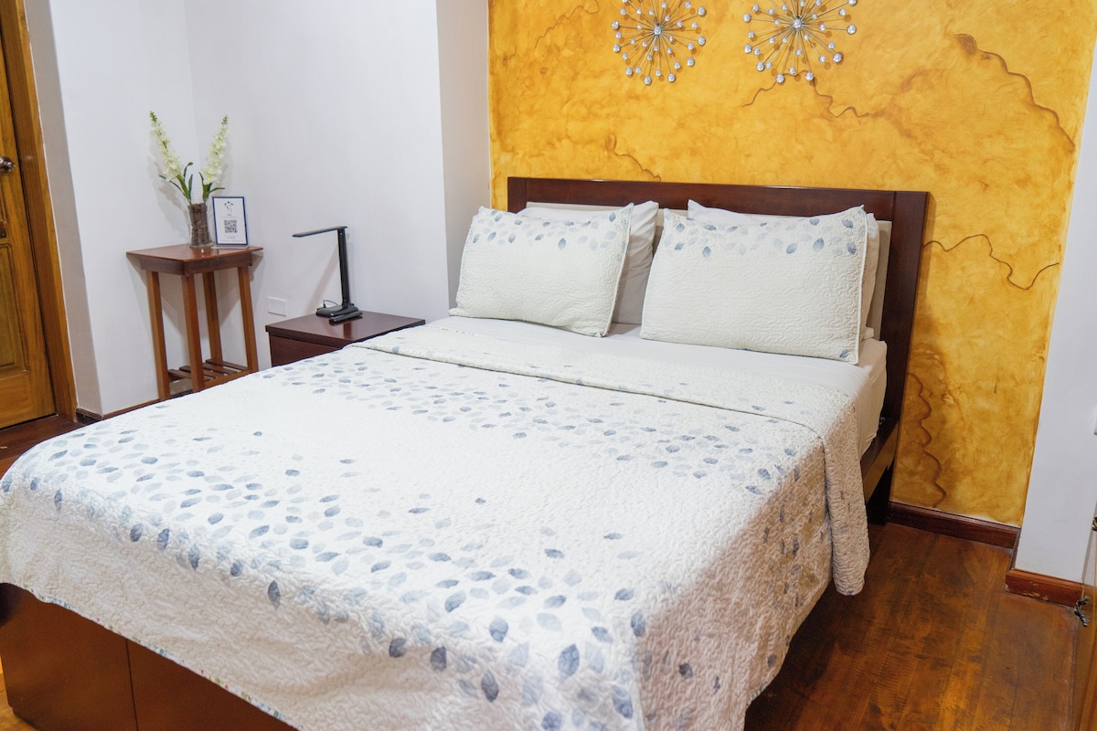 Quarto com cama de madeira, roupa de cama branca, mesa de cabeceira pequena com abajur e um objeto decorativo na parede.