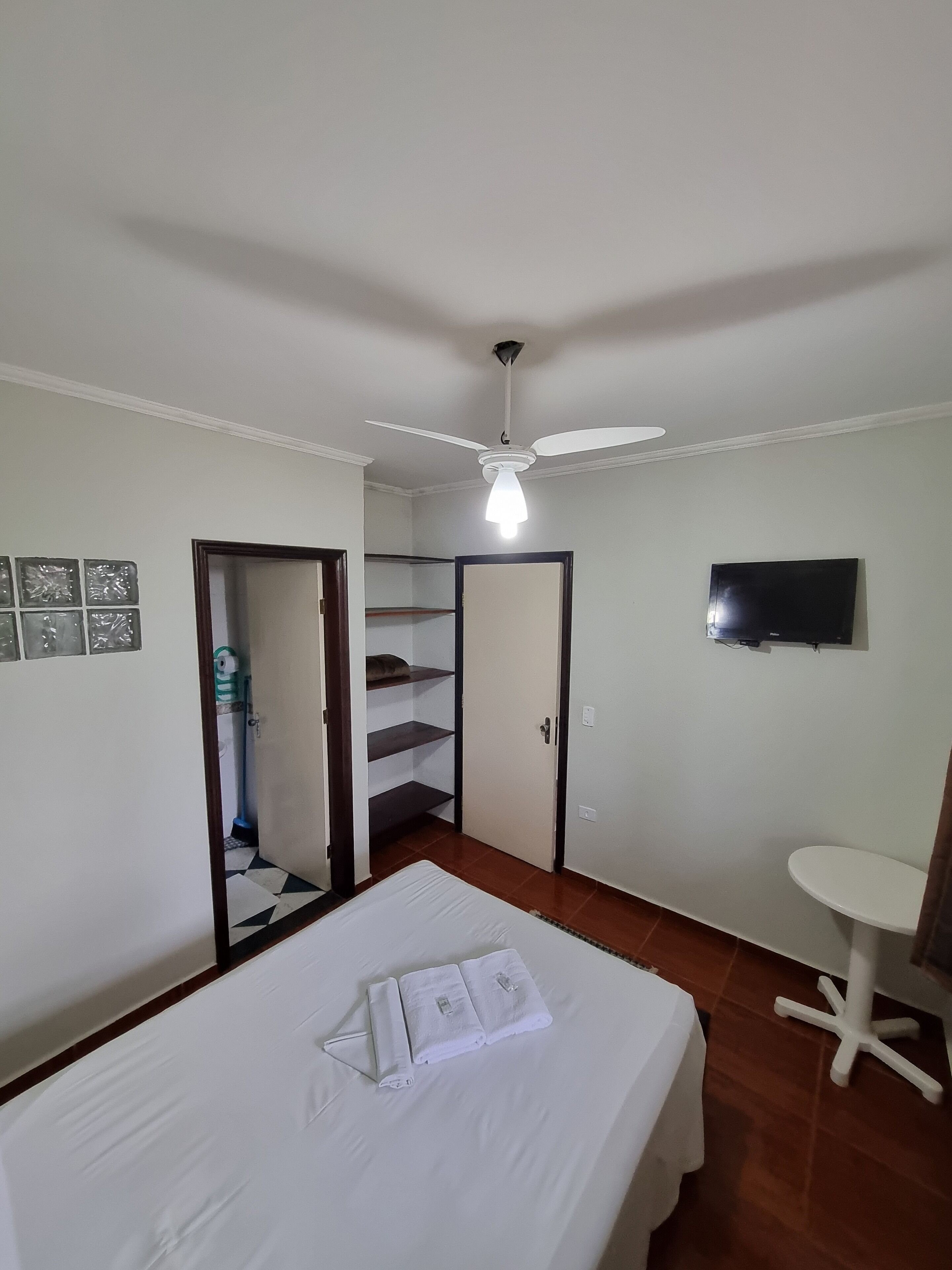 Quarto de hotel com cama, escrivaninha, cadeira, ventilador de teto e televisão fixada na parede.