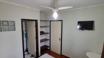 Quarto de hotel com cama, escrivaninha, cadeira, ventilador de teto e televisão fixada na parede.