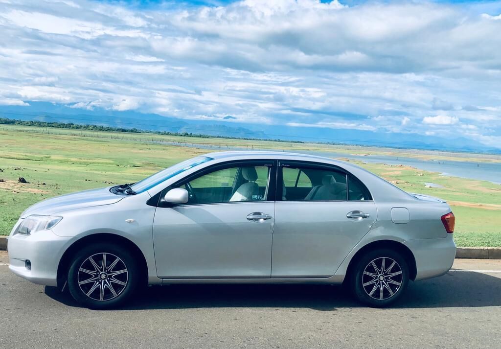 Um carro sedã branco estacionado em uma estrada, com vista panorâmica de um campo verde e um corpo d'água ao fundo.
