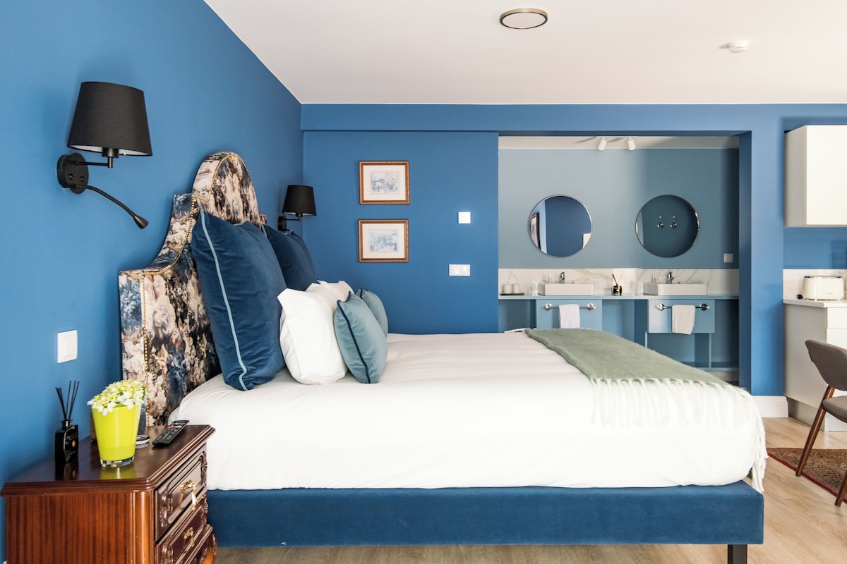 Quarto com uma cama grande, mesa de cabeceira, luminária, espelho e banheiro visível por uma porta aberta.