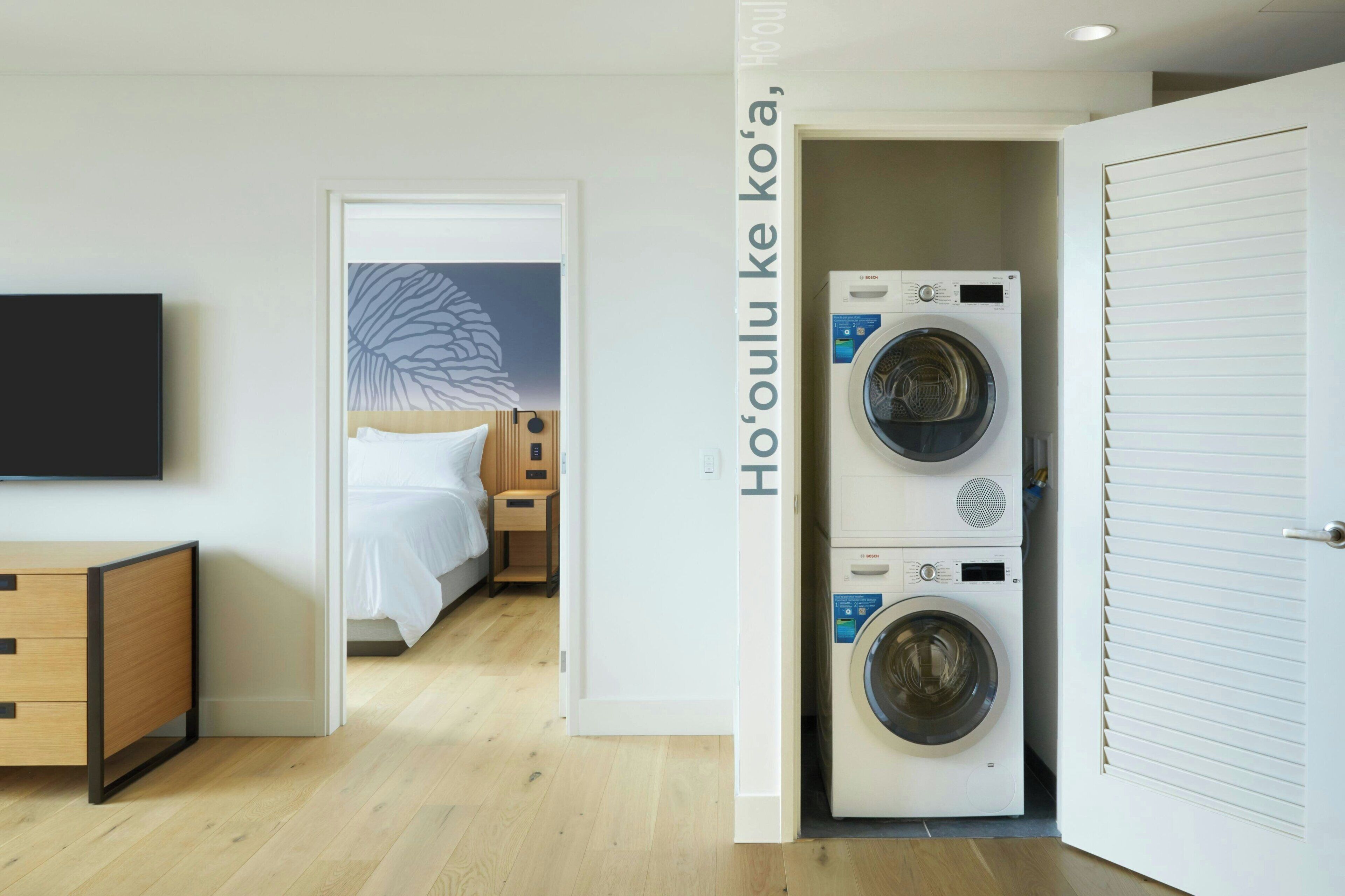 Quarto de hotel com cama, televisão, escrivaninha e máquina de lavar.