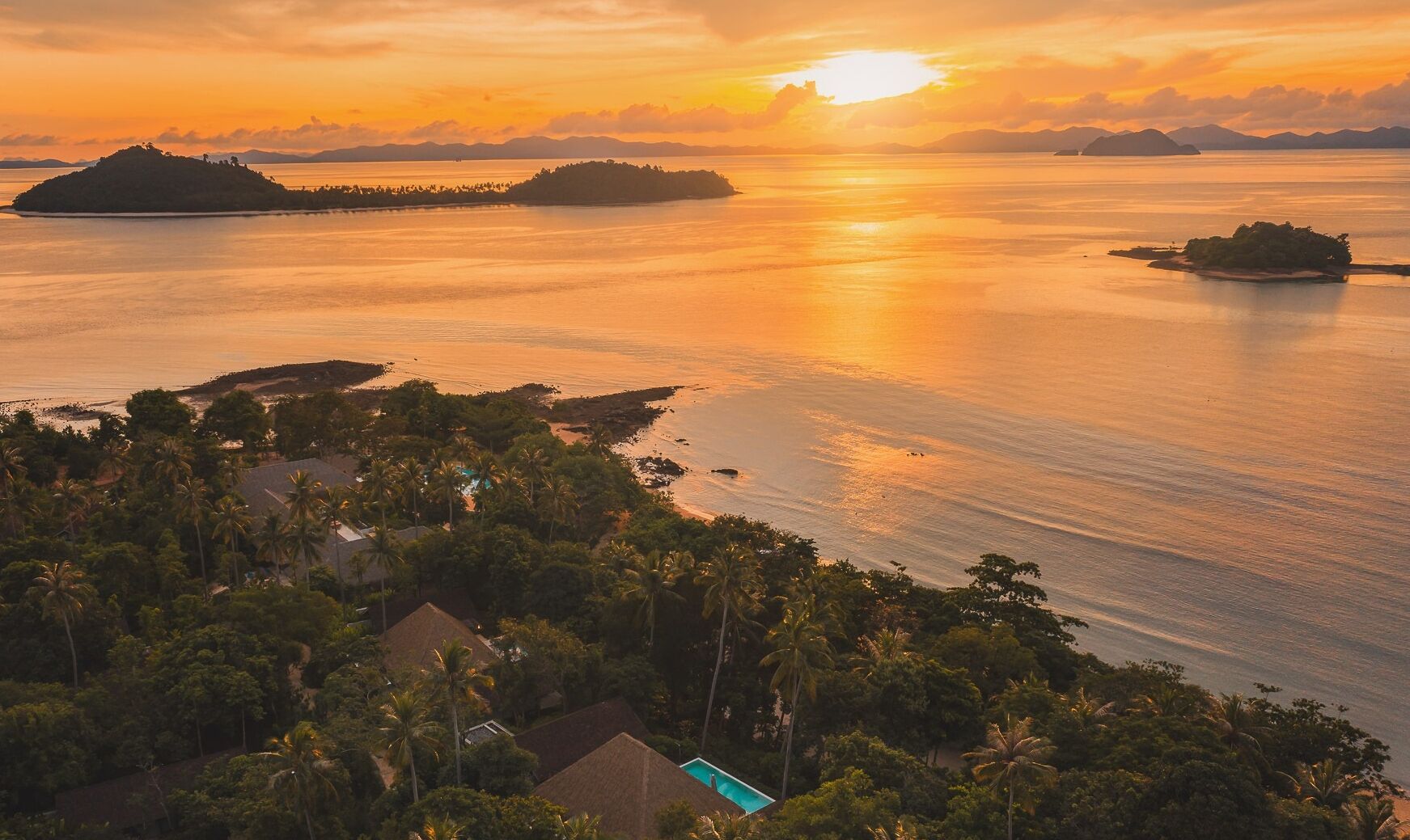 Resort tropical com piscina, cercado por vegetação exuberante e vista para um mar tranquilo com ilhas ao longe.