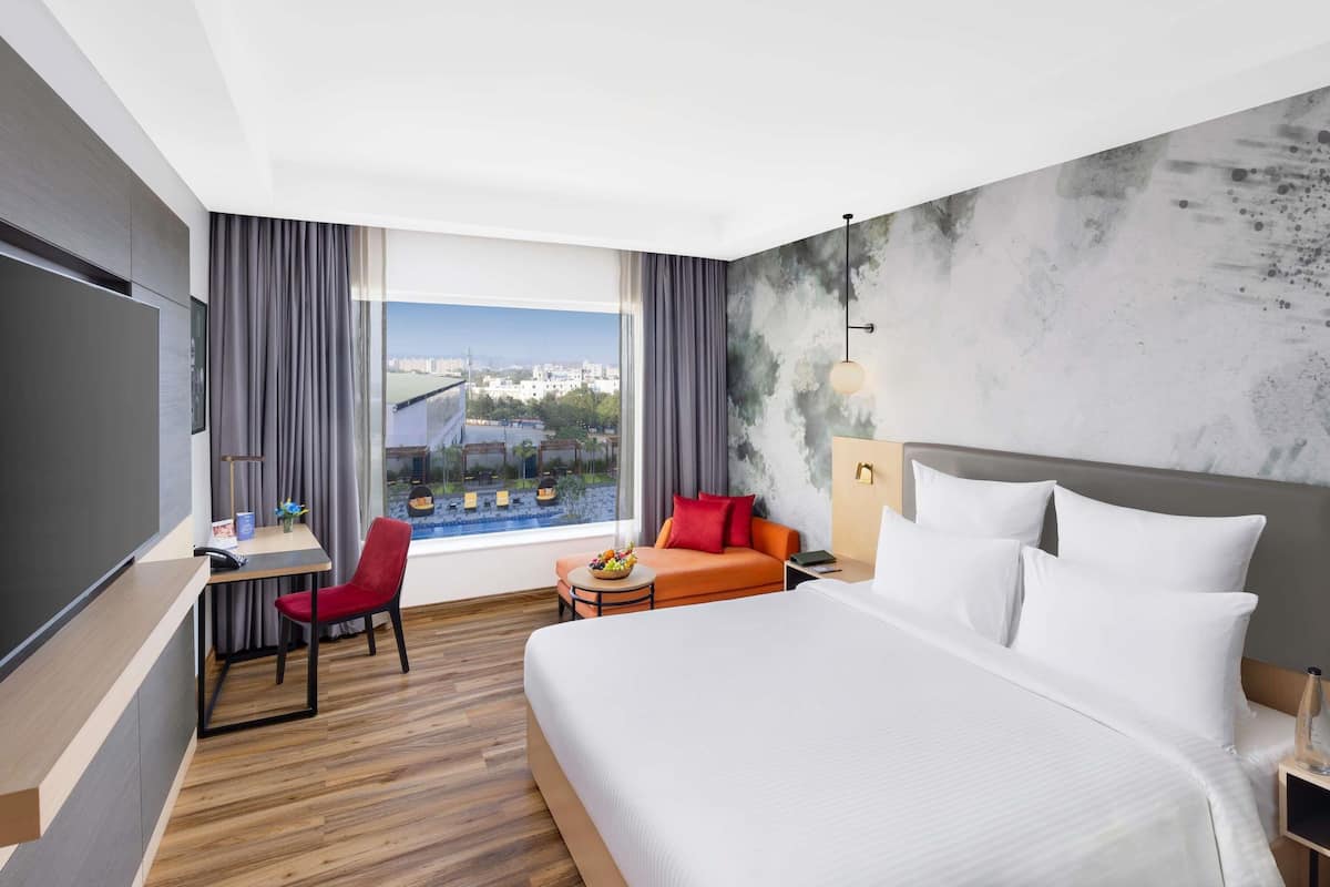 Quarto de hotel com cama, escrivaninha, cadeira, mesinha e vista para a piscina e prédios.