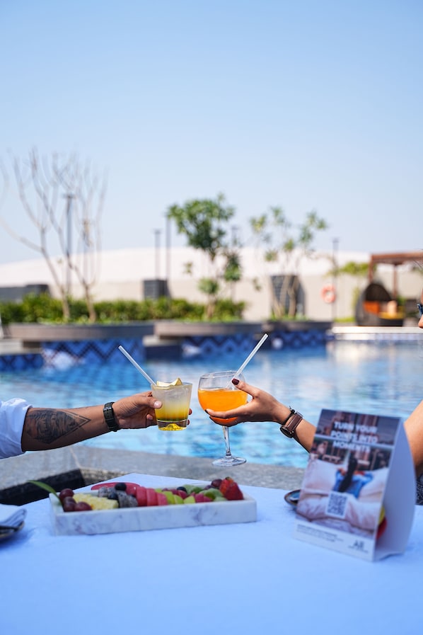 Duas pessoas brindando com bebidas em uma mesa à beira da piscina, com frutas e uma revista.