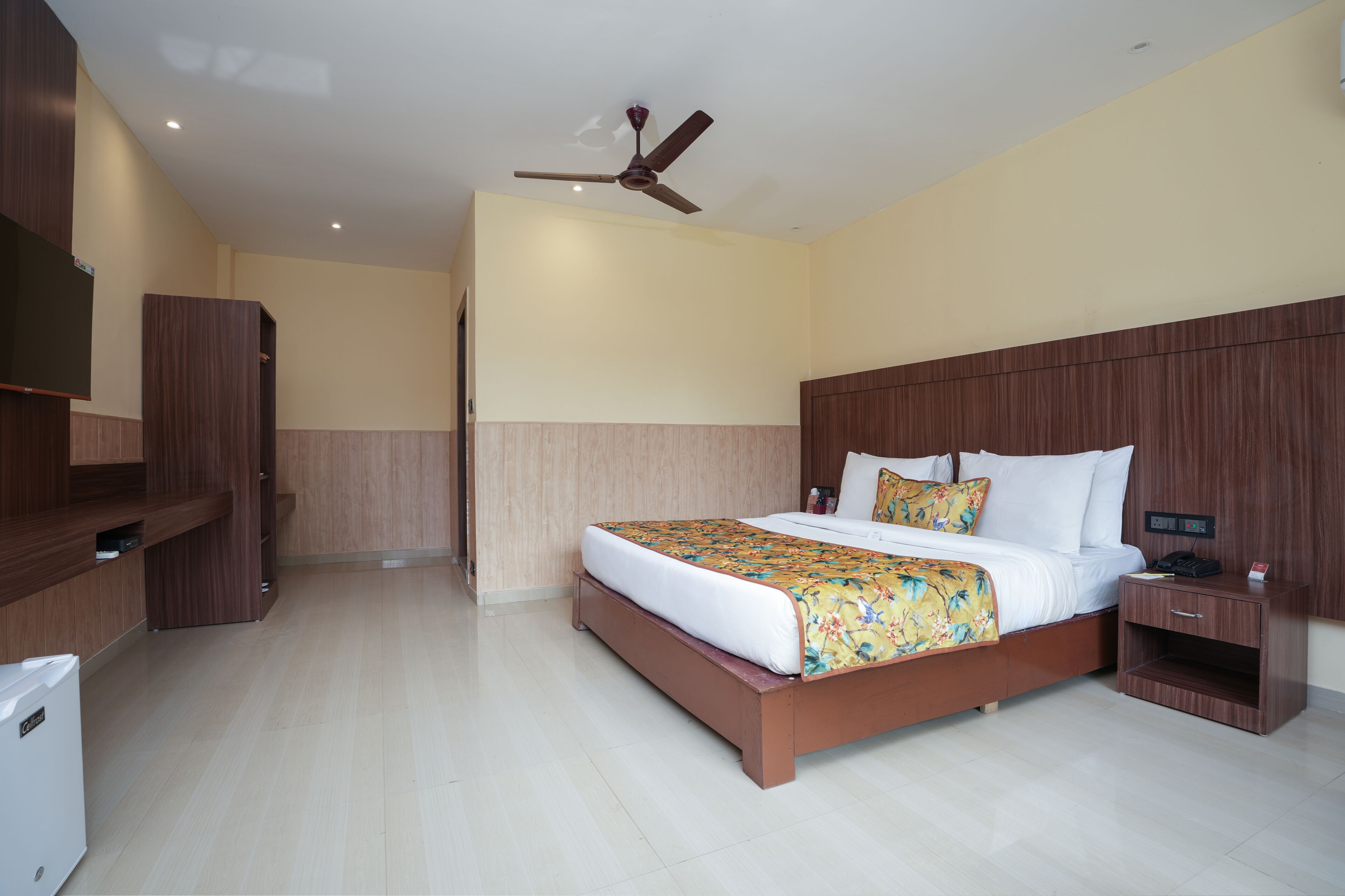 Quarto de hotel com cama, mesa de cabeceira, escrivaninha e ventilador de teto.