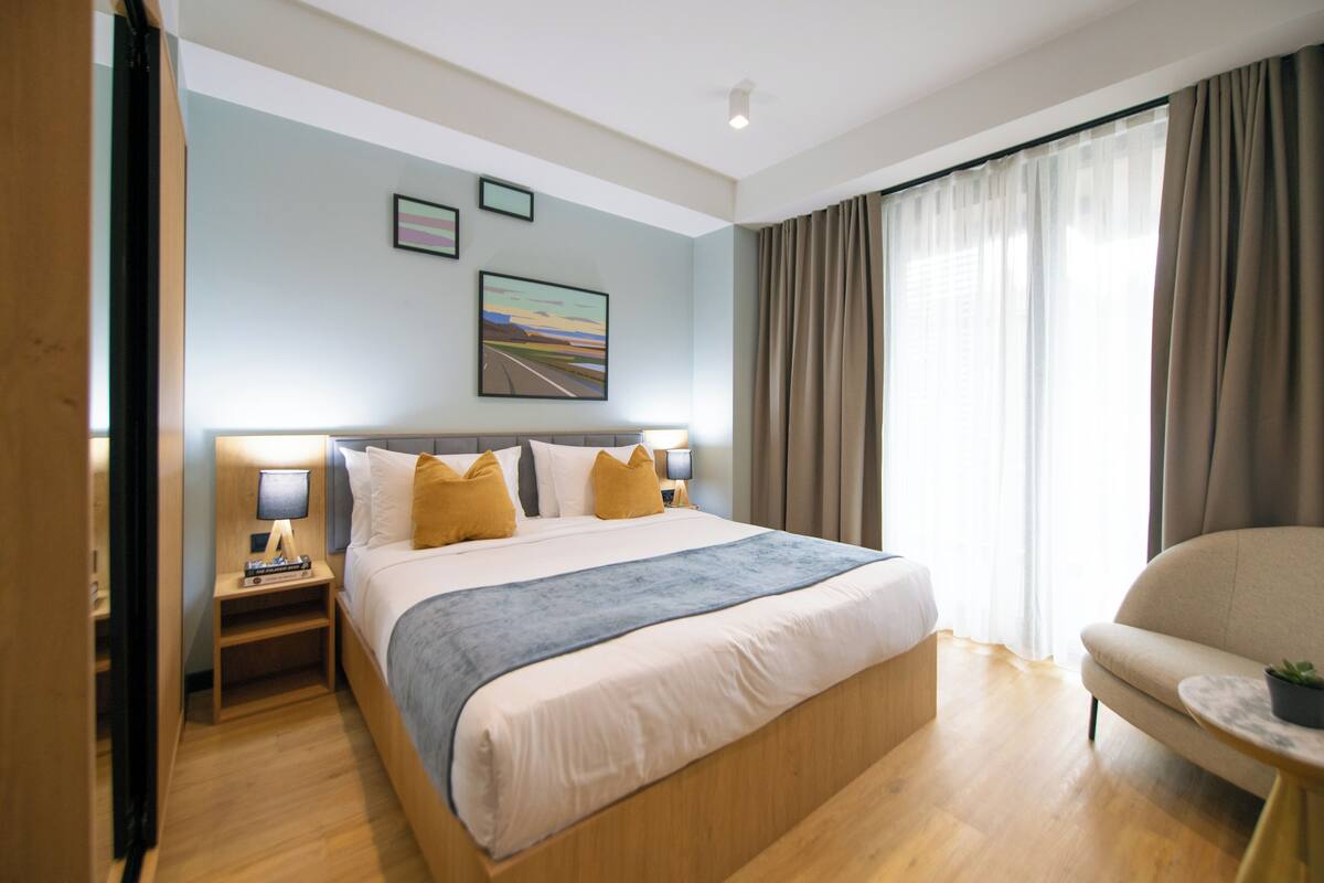 Quarto de hotel com uma cama grande, mesa de cabeceira com abajur, uma cadeira e janela com cortinas.