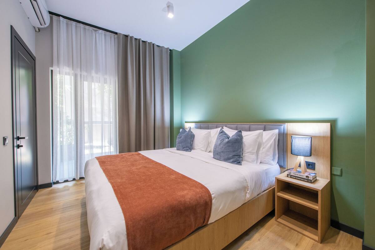 Quarto de hotel com uma cama grande, mesa de cabeceira com abajur e vista para o exterior através de cortinas leves.