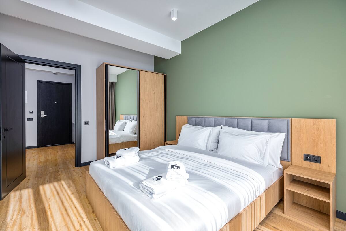 Quarto de hotel moderno com cama grande, piso de madeira, espelho e parede verde.