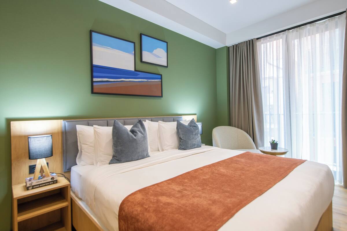 Quarto de hotel com uma cama grande, mesa de cabeceira com abajur, uma cadeira e uma pequena mesa com uma planta. Duas fotos emolduradas estão penduradas na parede acima da cama.