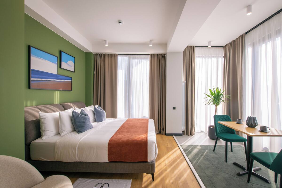Quarto de hotel moderno com uma cama grande, uma mesa e uma cadeira. Há cortinas nas janelas e uma planta na mesa.