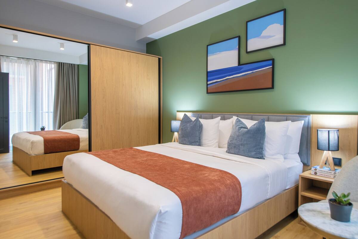 Quarto de hotel moderno com uma cama grande, um guarda-roupa, mesa de cabeceira com abajur e obras de arte na parede.