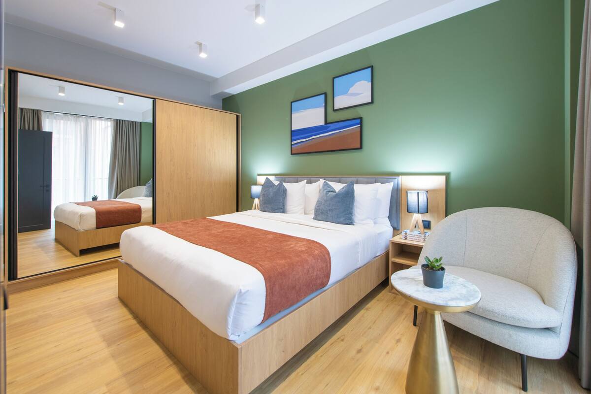 Quarto de hotel moderno com uma cama grande, mesa de cabeceira com luminária, uma cadeira e uma pequena mesa com uma planta.