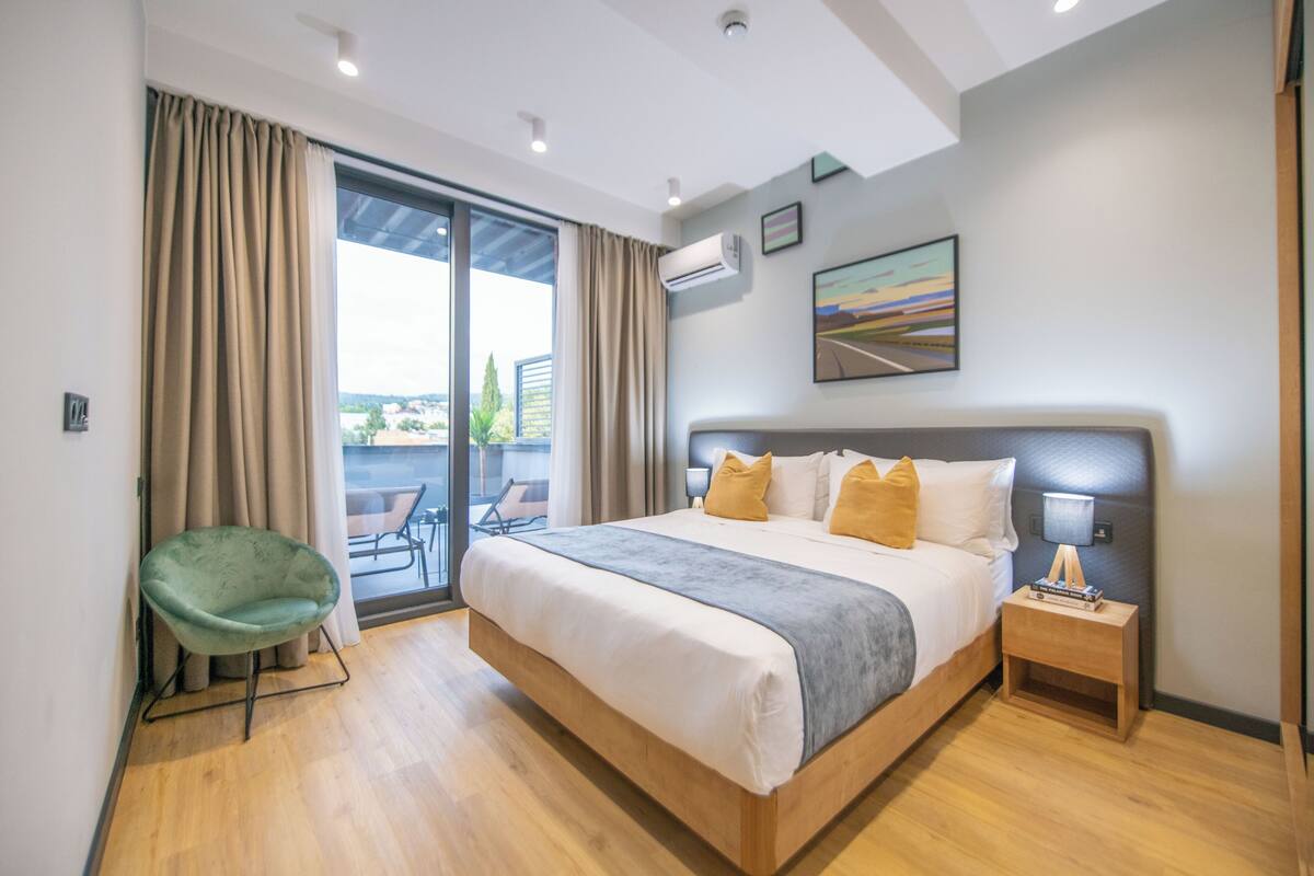 Quarto de hotel moderno com uma cama grande, uma cadeira verde, uma pequena mesa de cabeceira e vista para o exterior através de uma porta de vidro deslizante.