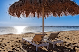 Uma praia com duas cadeiras de praia sob um toldo de palha, um cais ao fundo e céu limpo.