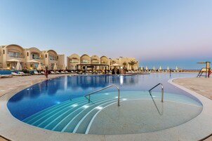 Piscina com design curvo, rodeada por espreguiçadeiras e guarda-sóis, com um edifício de hotel ao fundo.