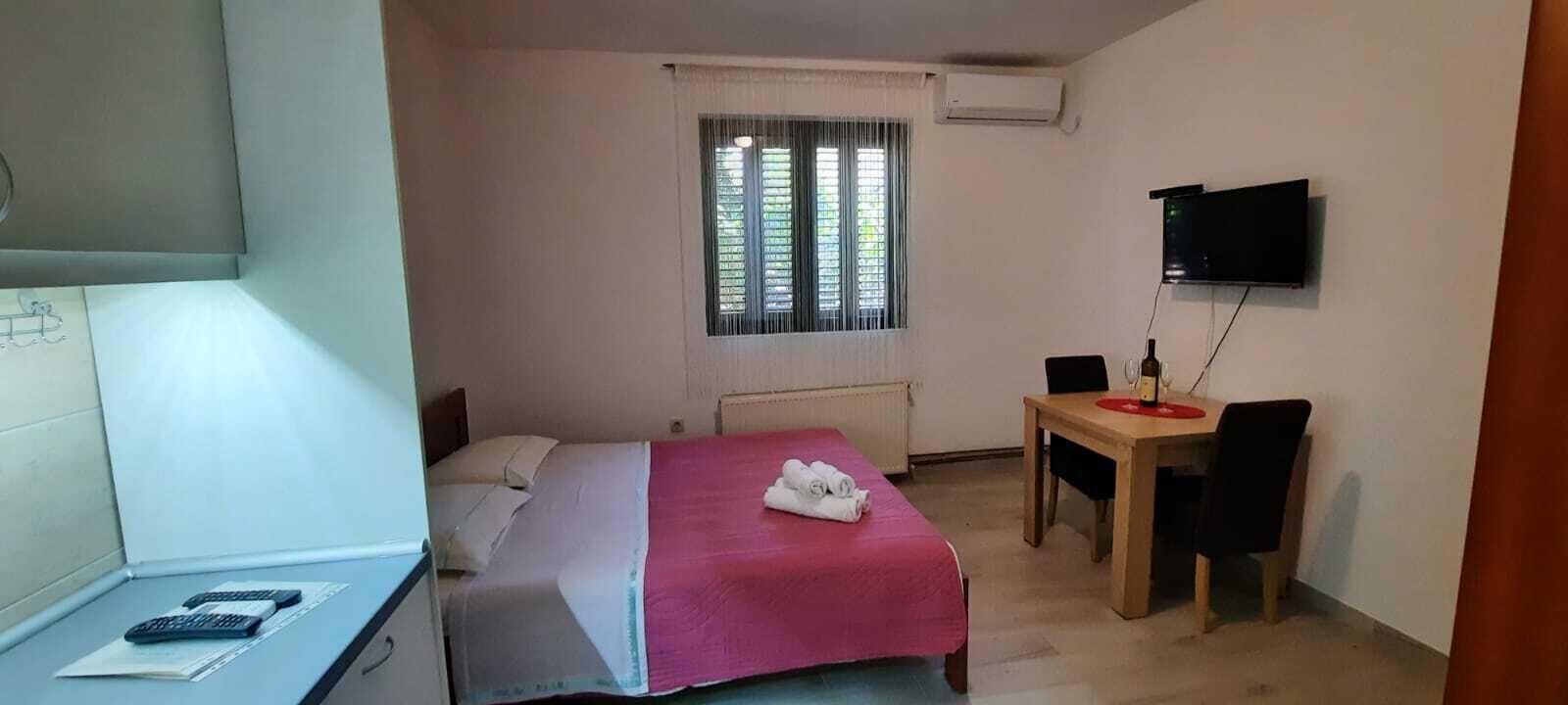 Quarto pequeno e limpo com uma cama, uma mesa com cadeira, uma TV fixada na parede e uma janela com persianas.