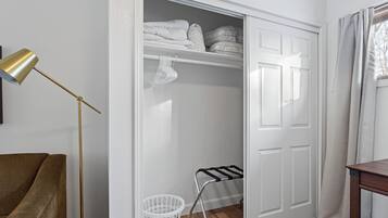 Um quarto com um guarda-roupa fechado, uma cadeira marrom, um candeeiro de chão dourado e uma janela com cortinas.