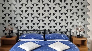 Um quarto com cabeceira estampada com padrão geométrico, cama com roupa de cama azul e branca com estrelas e duas luminárias laterais.