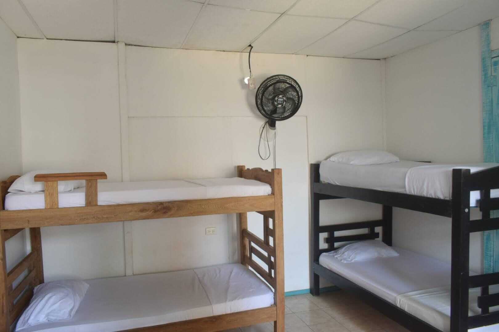 Quarto em dormitório com duas beliches, uma de madeira e outra de metal, e um ventilador de teto.