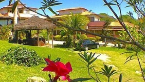 Um jardim tropical com um gazebo de palha, palmeiras e uma flor vermelha em primeiro plano.