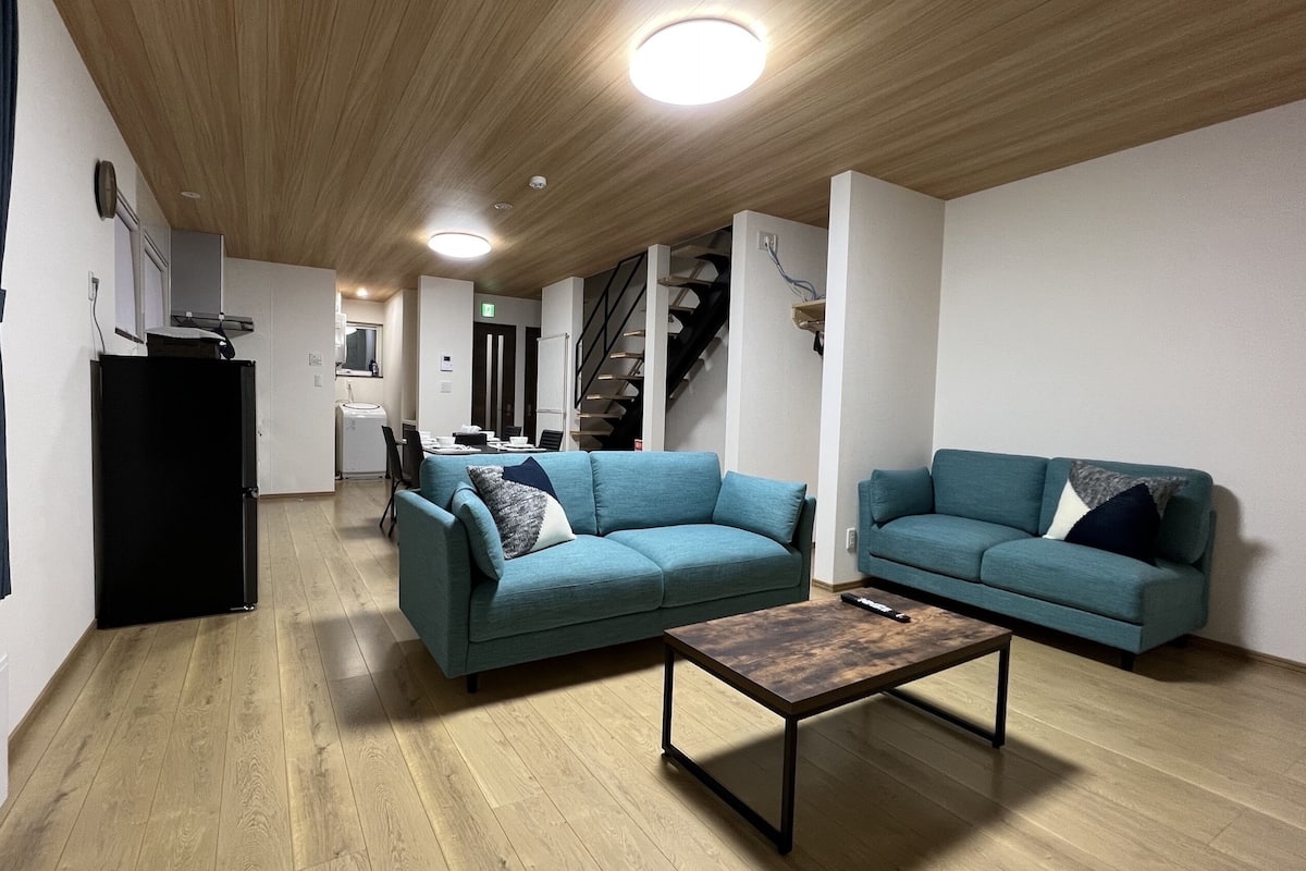 Uma sala de estar moderna com dois sofás azul-turquesa, uma mesa de centro de madeira e uma cozinha ao fundo.