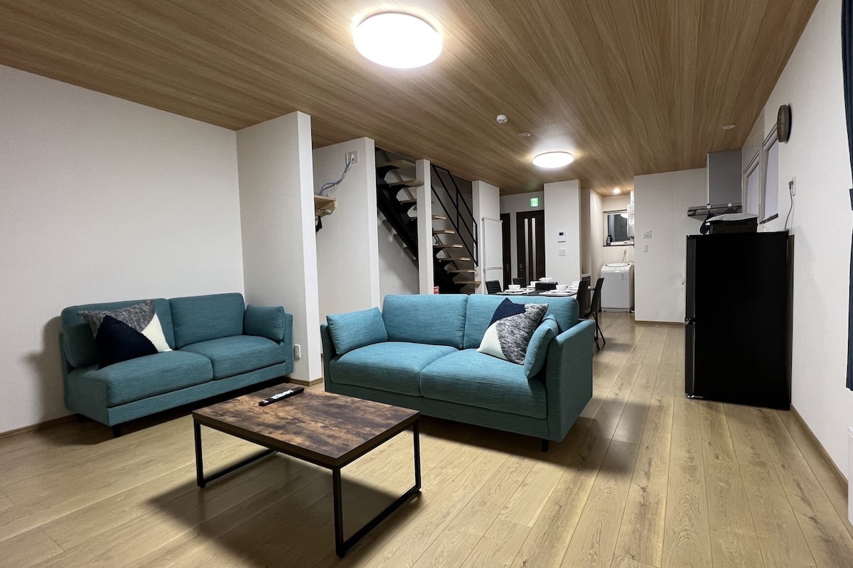 Uma sala de estar moderna com dois sofás verde-água, uma mesa de centro de madeira e uma escada que dá para uma área de cozinha.