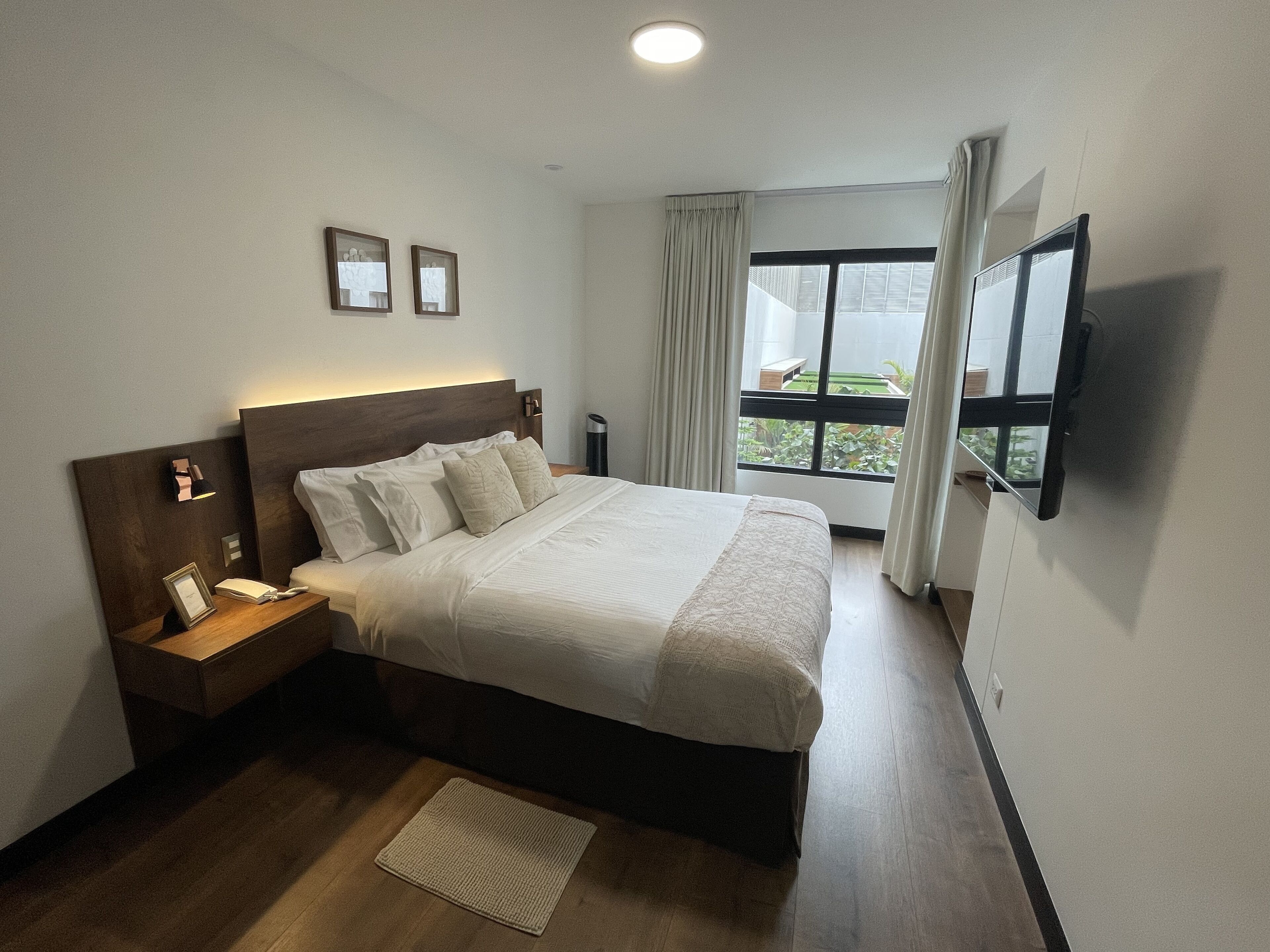 Quarto de hotel com cama, mesa de cabeceira, abajur e televisão na parede.
