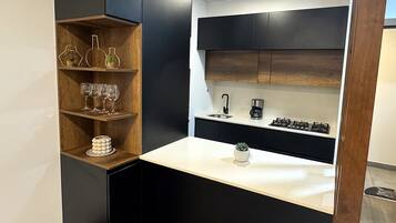 Uma cozinha compacta com armários escuros, bancada branca e uma prateleira com objetos decorativos.