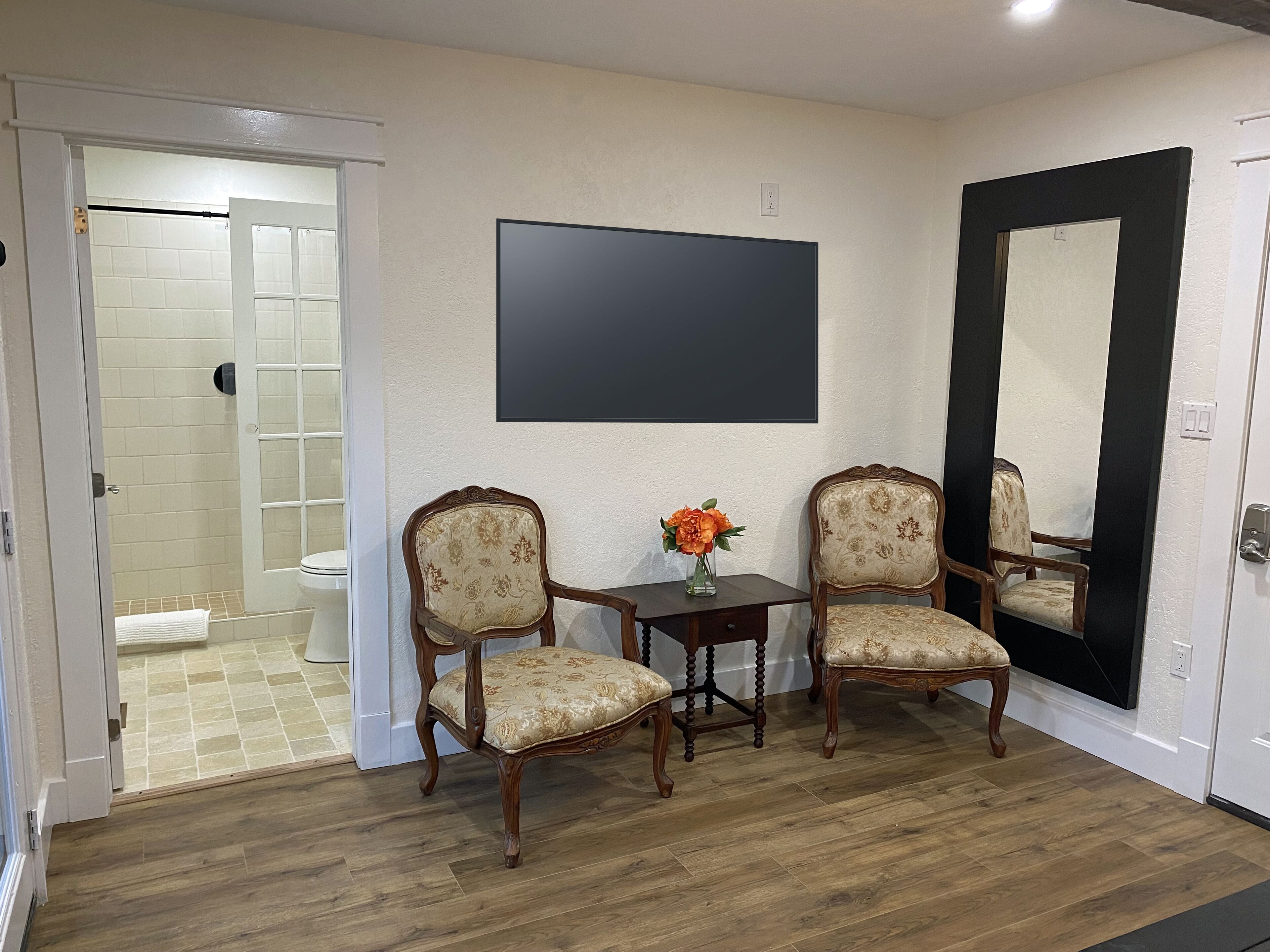 Um quarto com duas cadeiras decorativas, uma mesa pequena com um vaso e um espelho grande na parede.