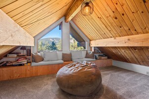 Um quarto de sótão aconchegante com vigas de madeira, um grande puff e vista para uma encosta arborizada.