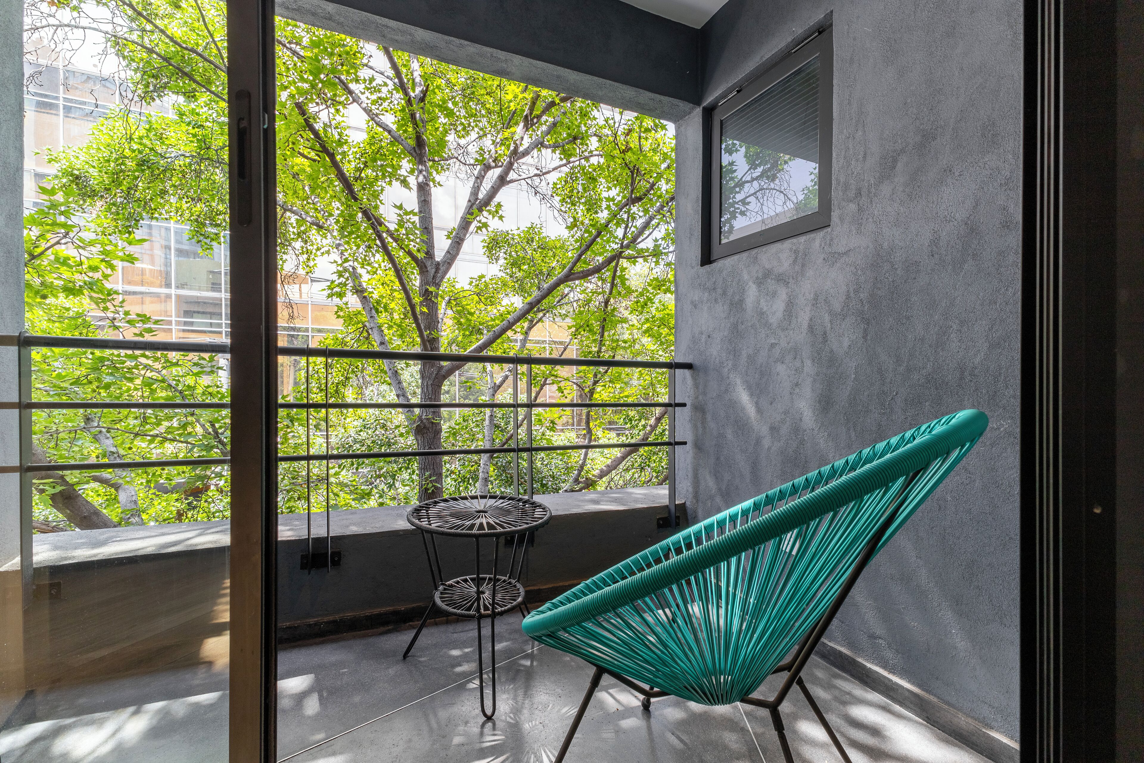 Uma varanda com uma cadeira verde, uma mesa pequena e vista para árvores e edifícios.