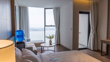 Quarto de hotel moderno com uma cama grande, um sofá, uma mesa pequena e vista para o mar por uma janela ampla.