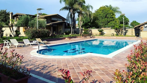 Piscina com deck ladrilhado, cercada por palmeiras e algumas casas ao fundo.