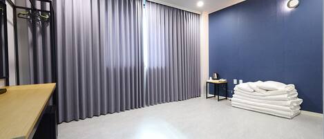 Quarto de hotel com uma grande janela coberta por cortinas cinzentas, uma escrivaninha de madeira, uma pequena mesa com abajur e uma pilha de toalhas brancas em frente a uma parede azul.