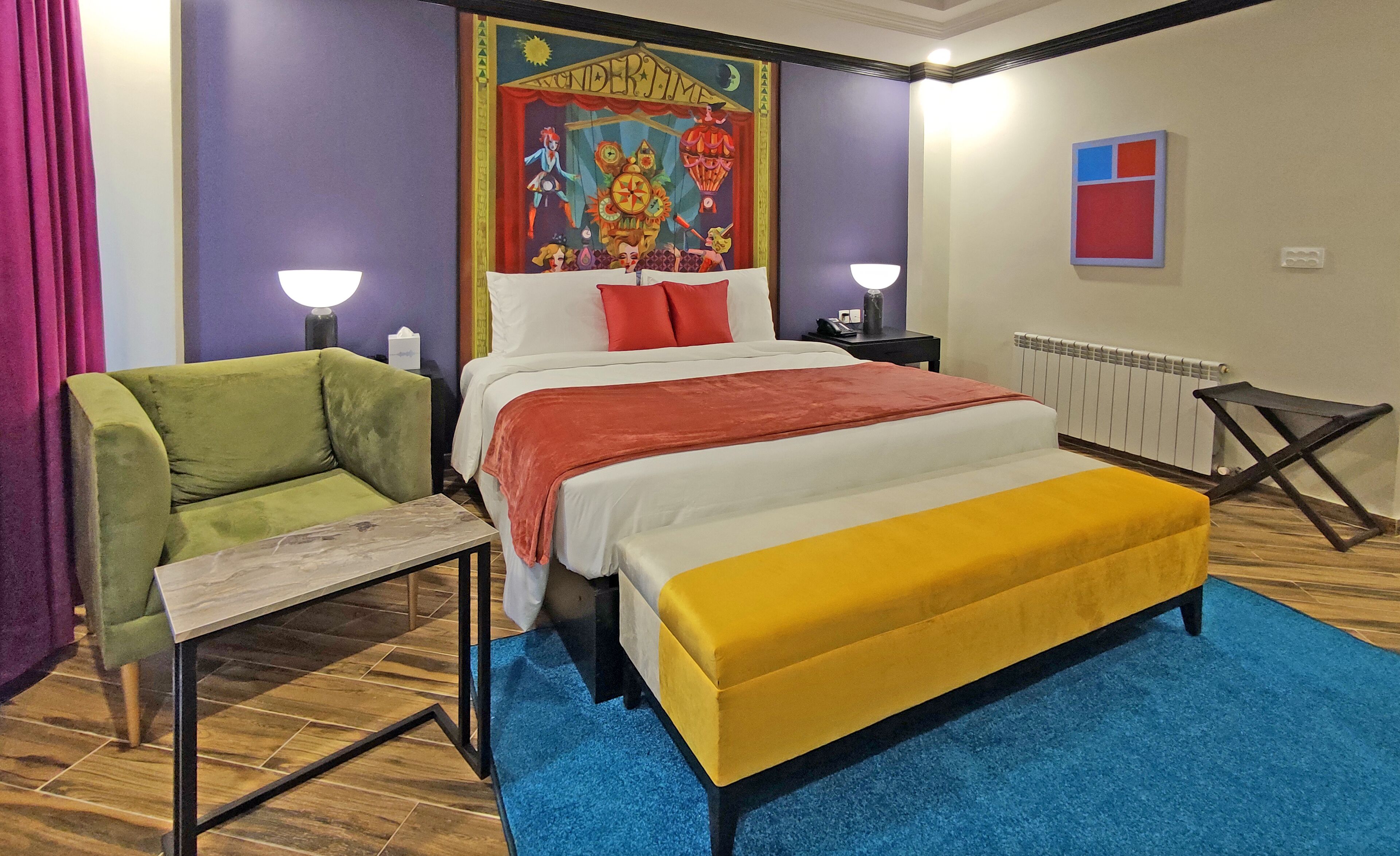 Quarto com cama, poltrona verde, mesa pequena, banco, painel de parede colorido e mesa de cabeceira com abajur.