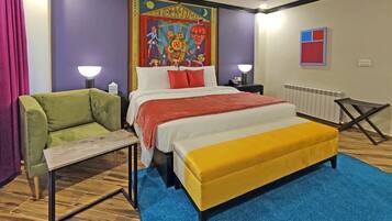 Quarto com cama, poltrona verde, mesa pequena, banco, painel de parede colorido e mesa de cabeceira com abajur.