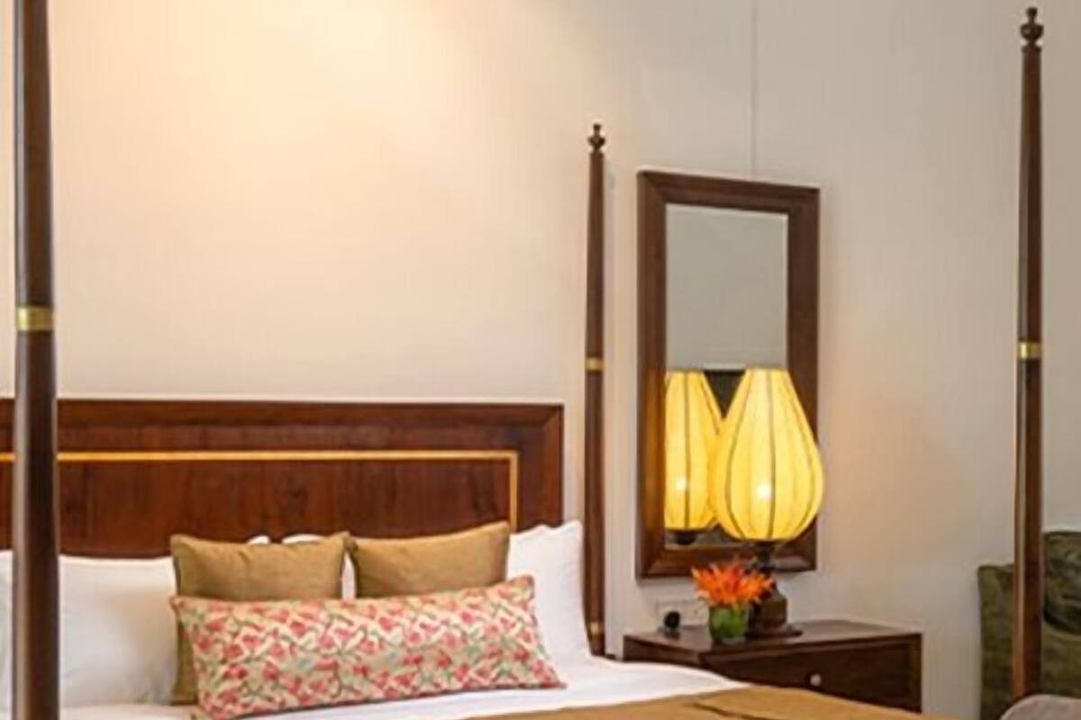 Uma cama de dossel com cabeceira de madeira, mesa de cabeceira com abajur e um banquinho estofado.