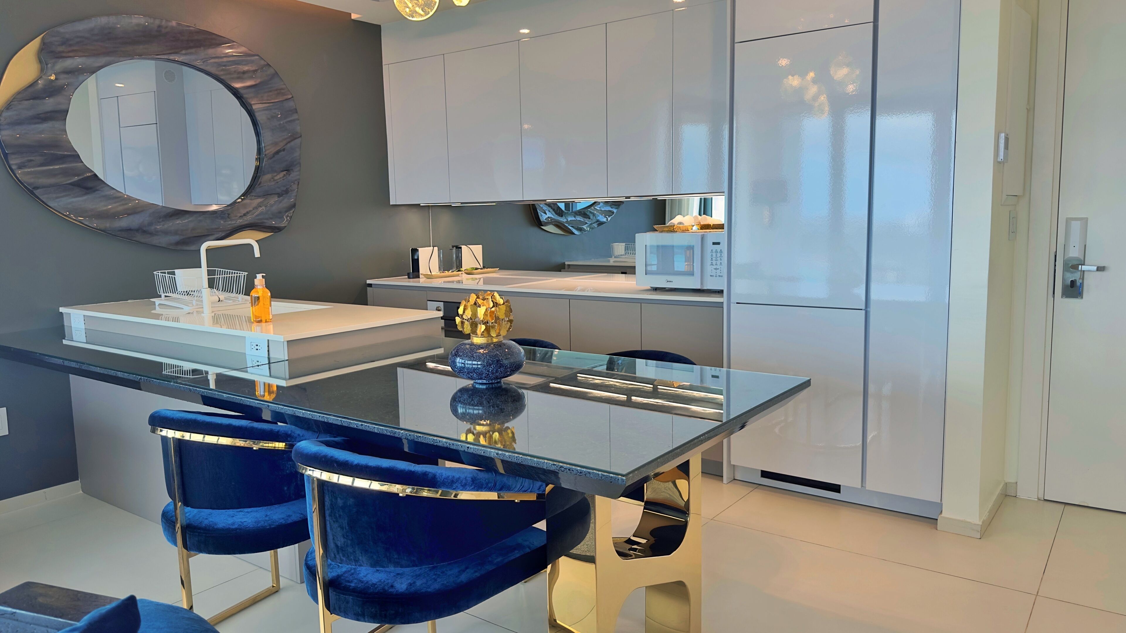 Uma cozinha moderna com uma mesa de jantar de vidro, duas cadeiras de veludo azul, um micro-ondas branco e um espelho grande na parede.