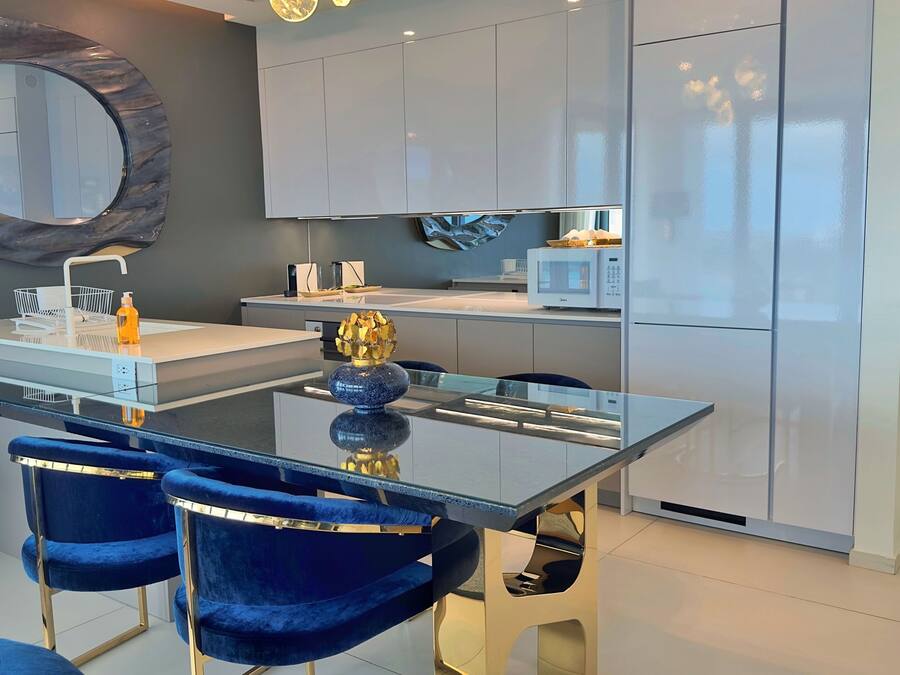 Uma cozinha moderna com uma mesa de jantar de vidro, duas cadeiras de veludo azul, um micro-ondas branco e um espelho grande na parede.