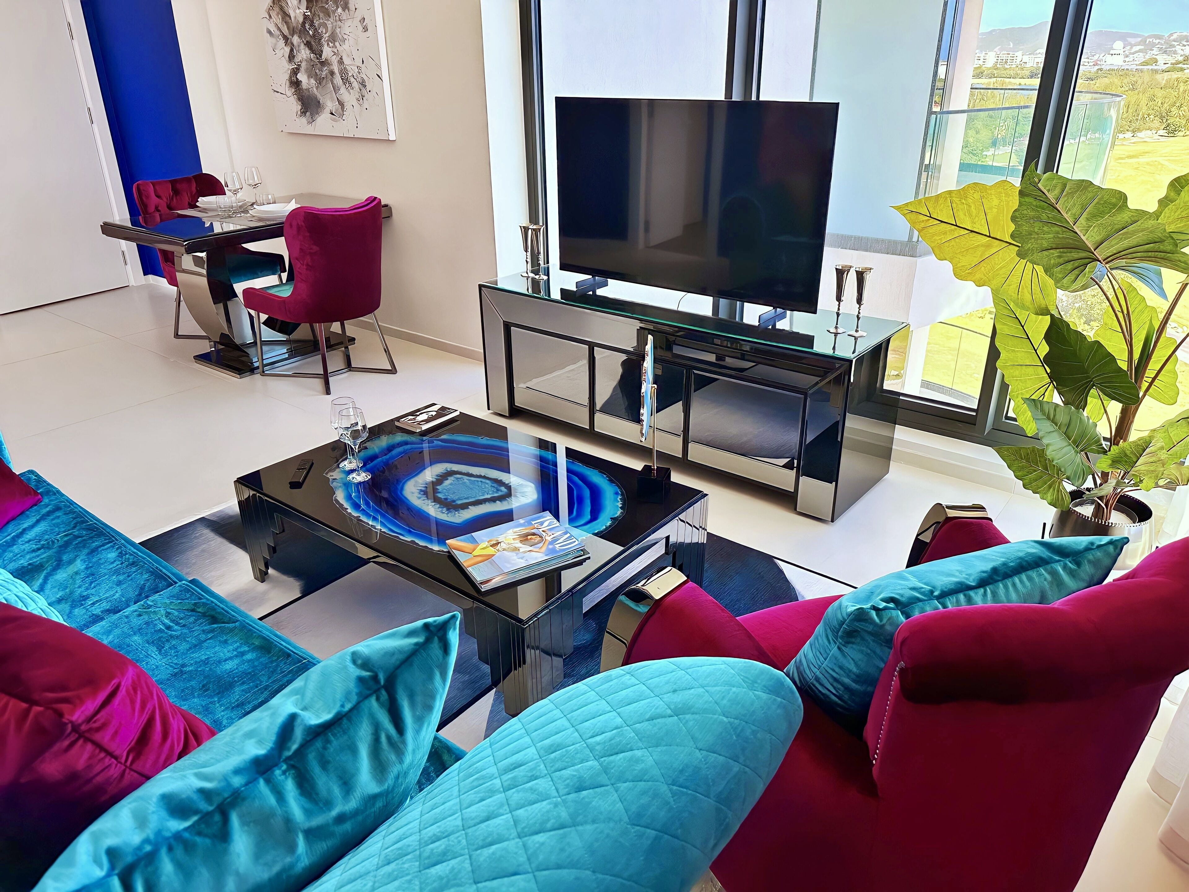Uma sala de estar moderna com uma televisão de tela plana grande, uma mesa de centro de vidro com um objeto decorativo azul e uma planta grande.