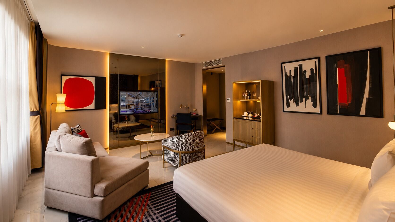 Quarto de hotel com cama, sofá, televisão, escrivaninha e obras de arte nas paredes.