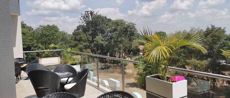 Varanda com mobiliário de vime, guarda-corpo de vidro e vista para árvores e um edifício ao fundo.