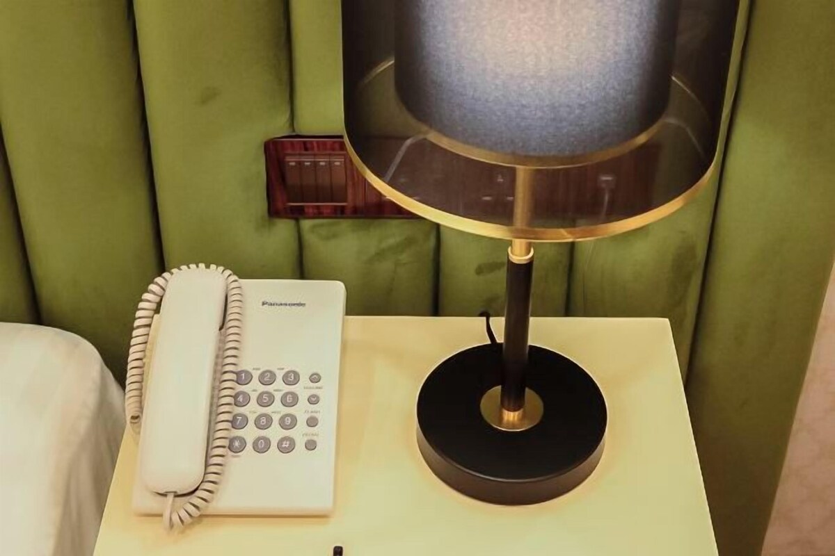 Quarto com cabeceira estofada em verde, telefone branco e abajur preto.