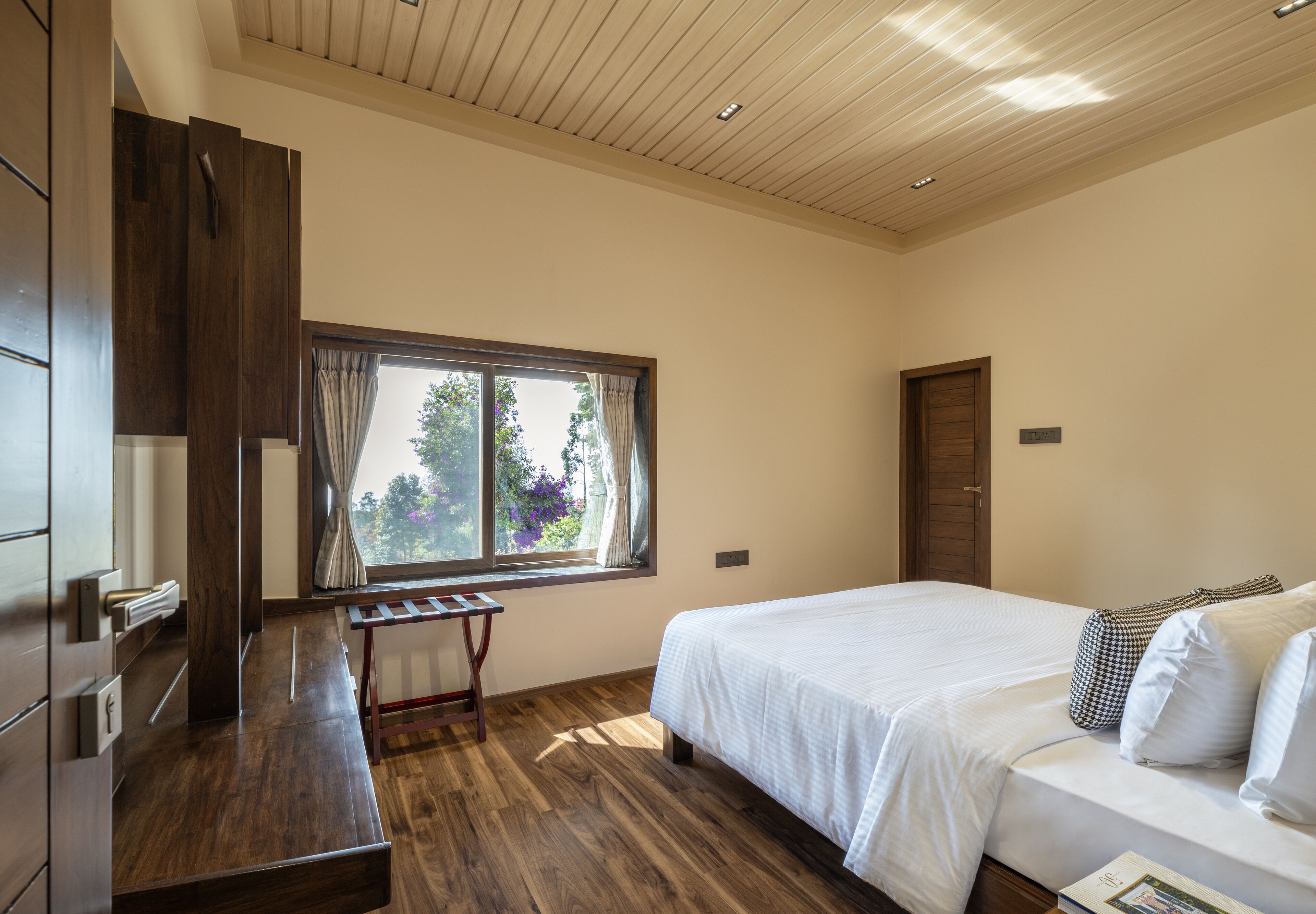 Quarto com piso de madeira, cama com roupa de cama branca, escrivaninha com cadeira e janela com cortinas.