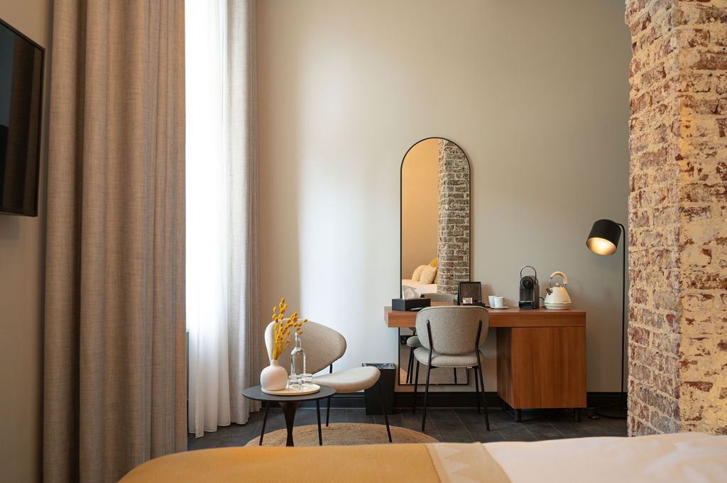 Quarto de hotel com uma cama, uma escrivaninha com cadeira, um espelho grande e um abajur.