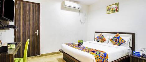 Quarto de hotel com cama, escrivaninha, cadeira, ar condicionado, ventoinha no teto e um quadro na parede.